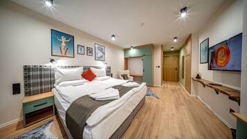 Quarto de hotel moderno com uma cama grande, mesa de cabeceira, chão de madeira e corredor que leva a uma porta.