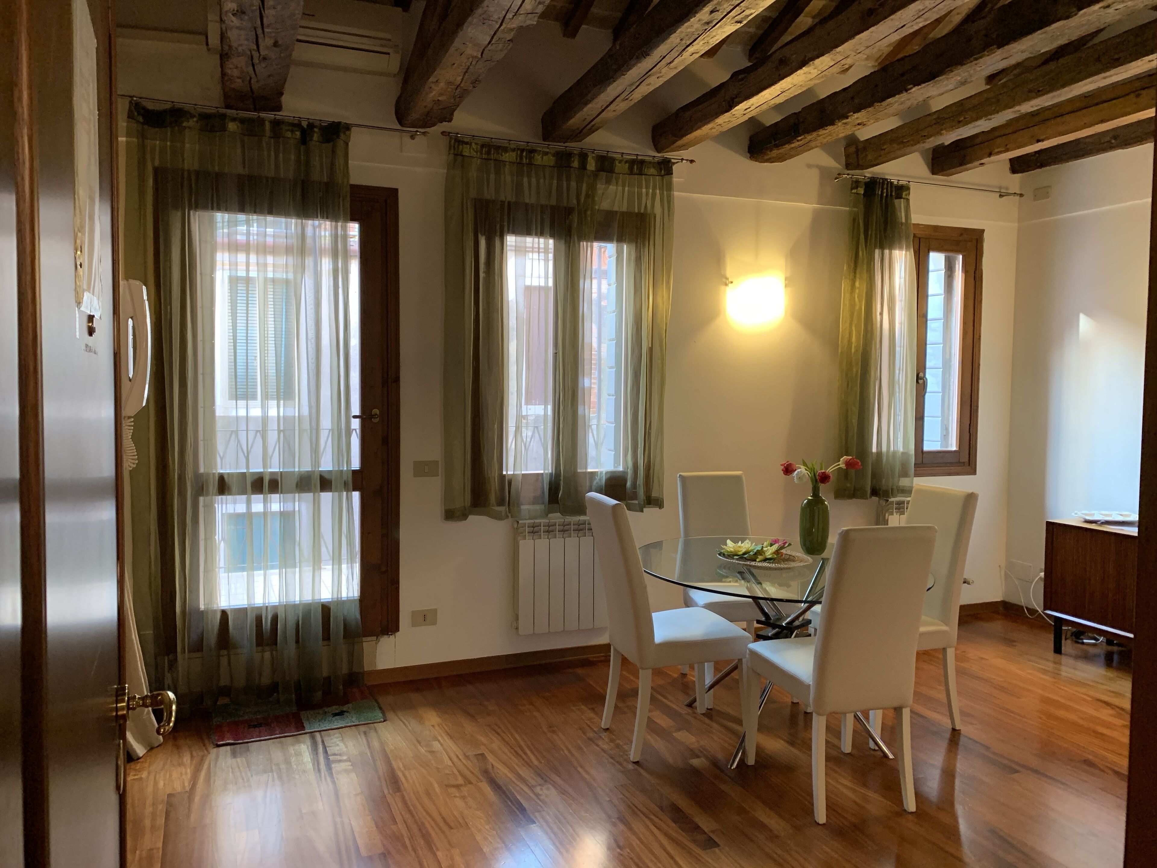 Uma sala de jantar com mesa de vidro, cadeiras brancas e um vaso com flores.