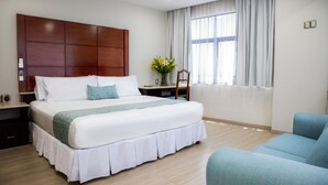 Quarto de hotel com uma cama grande, uma mesa de trabalho com cadeira, janela com cortinas e ventoinha no teto.