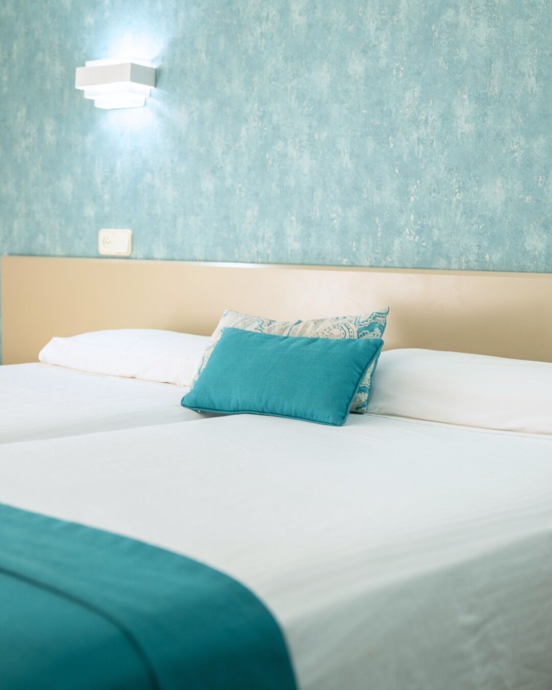 Quarto de hotel com duas camas, parede azul clara com textura e luminária fixa na parede.