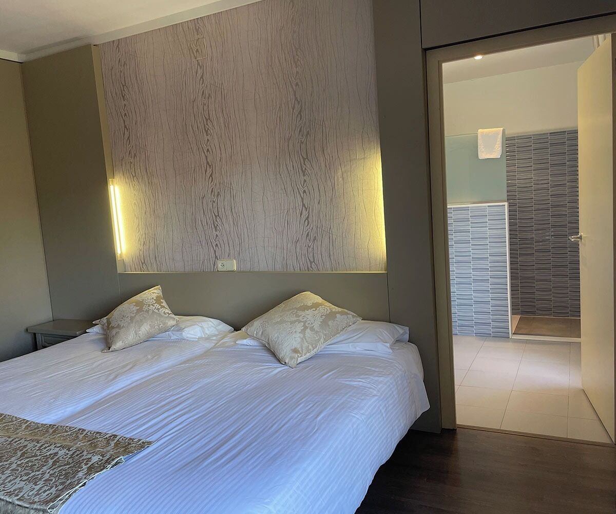 Quarto de hotel moderno com uma cama grande, duas almofadas, uma mesa de cabeceira e um aplique de parede.