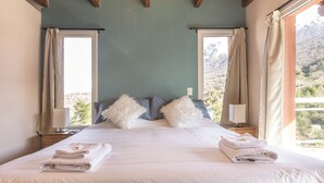 Cama bem arrumada com lençóis brancos e almofadas fofas, mesa de cabeceira com abajur e janela com cortinas.
