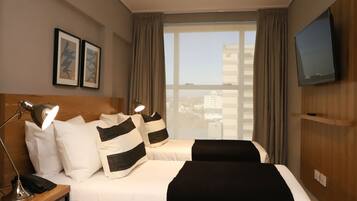 Quarto de hotel com duas camas, uma TV, escrivaninha com luminária e uma janela grande com cortinas.