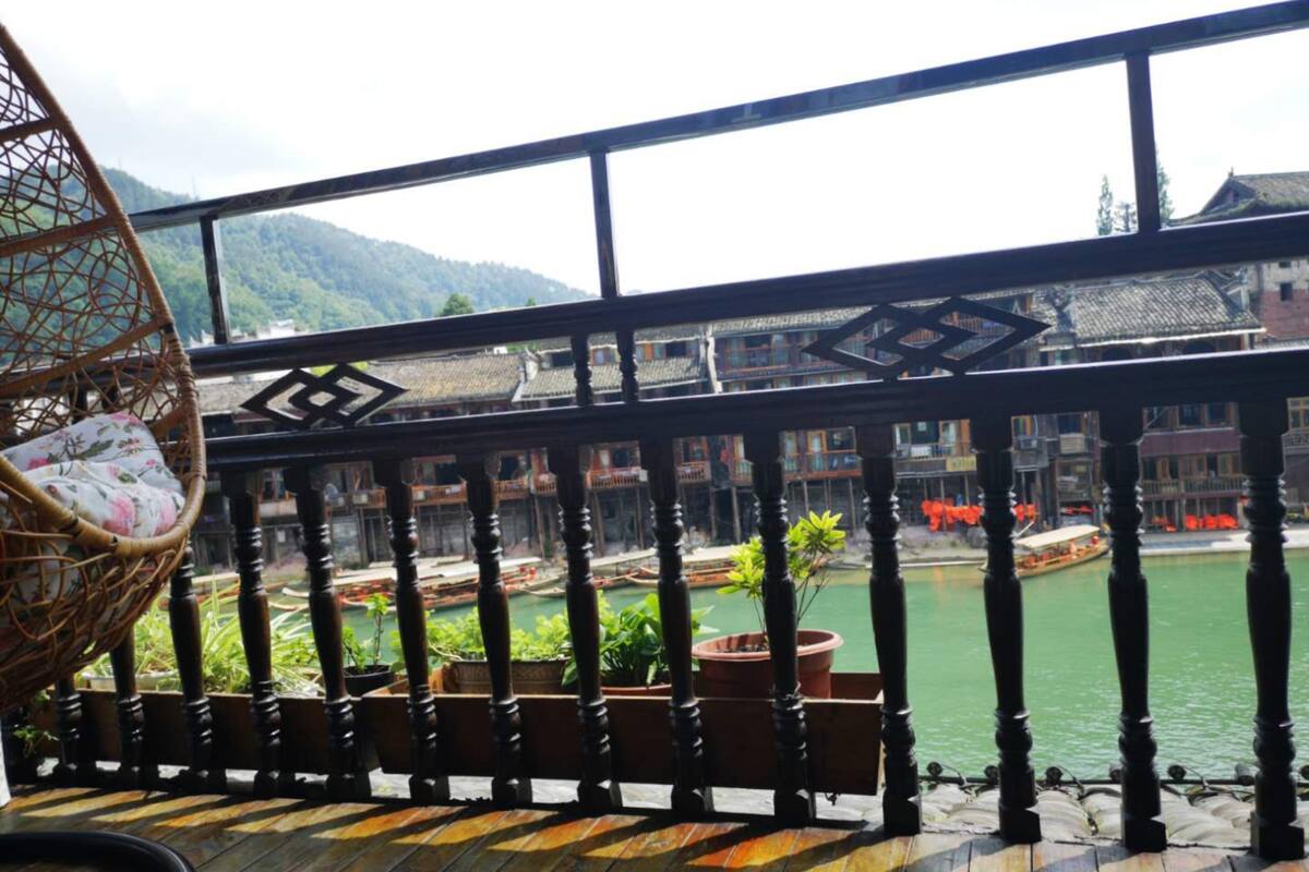 Um deck de madeira com uma cadeira de vime, plantas em vasos e vista para edifícios tradicionais à beira do rio.