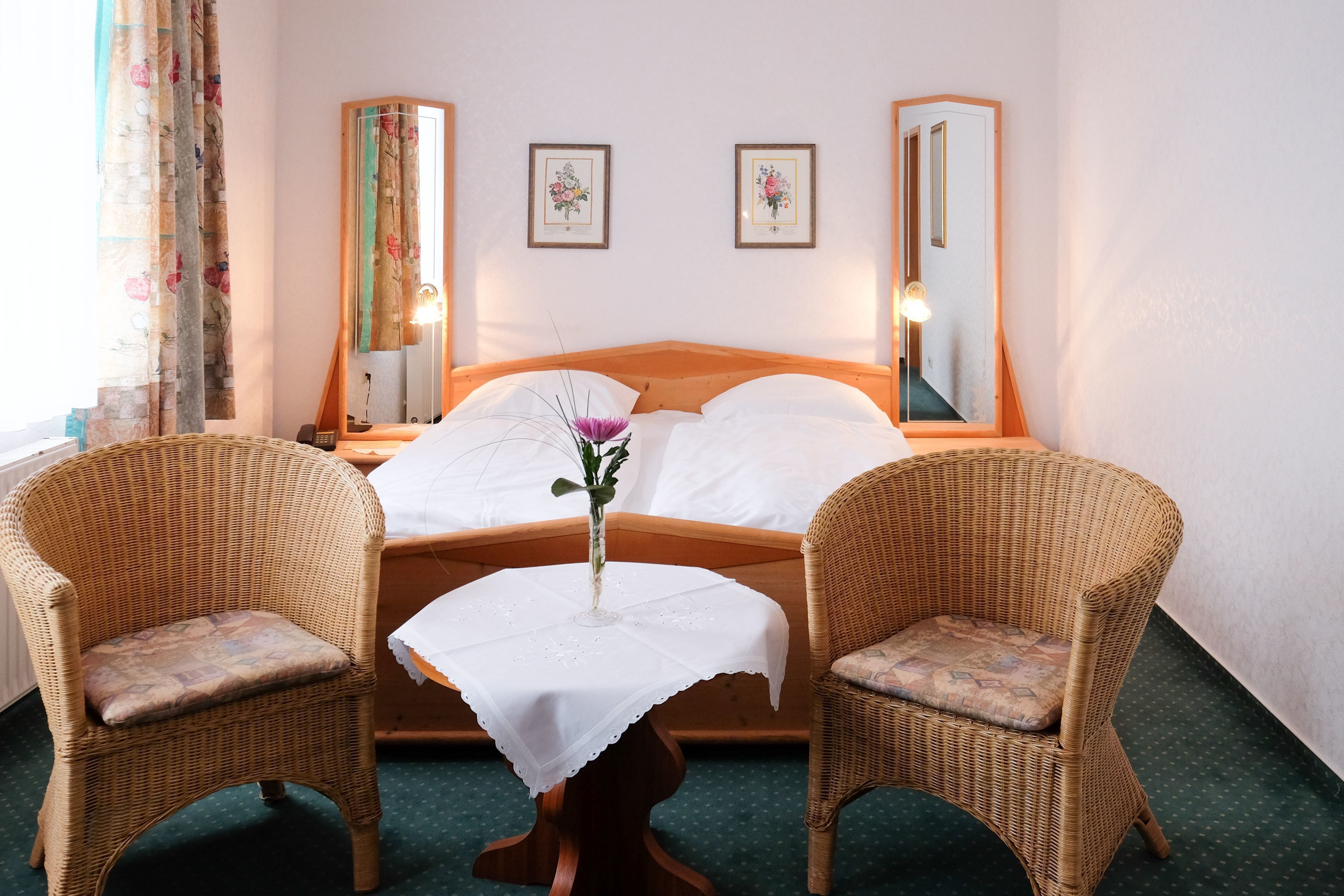 Quarto de hotel com cama, duas cadeiras de vime, uma mesinha com um vaso e duas quadros decorativos com flores na parede.