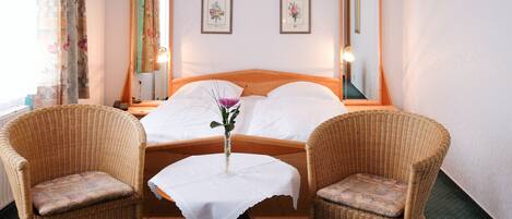 Quarto de hotel com cama, duas cadeiras de vime, uma mesinha com um vaso e duas quadros decorativos com flores na parede.