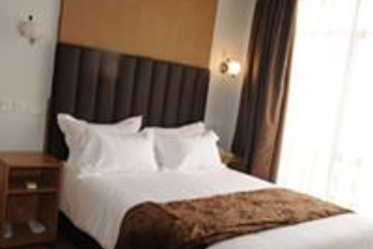 Quarto de hotel com uma cama grande, cabeceira de madeira, mesa de cabeceira e uma janela com cortinas.