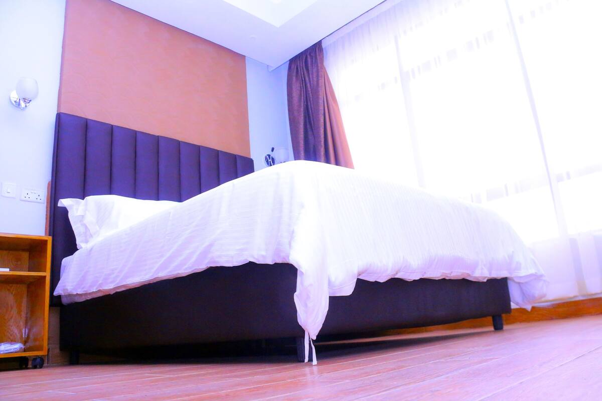 Quarto de hotel com cama, piso de madeira, prateleira na parede e janela com cortinas.