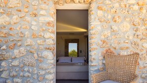 Quarto com paredes de pedra, uma cama, uma cadeira de vime e vista através de uma porta aberta.