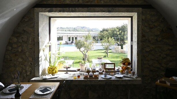 Uma área de refeições com parede de pedra, janela com vista para um edifício e um gramado, e uma mesa posta com pratos, talheres e uma seleção de frutas.