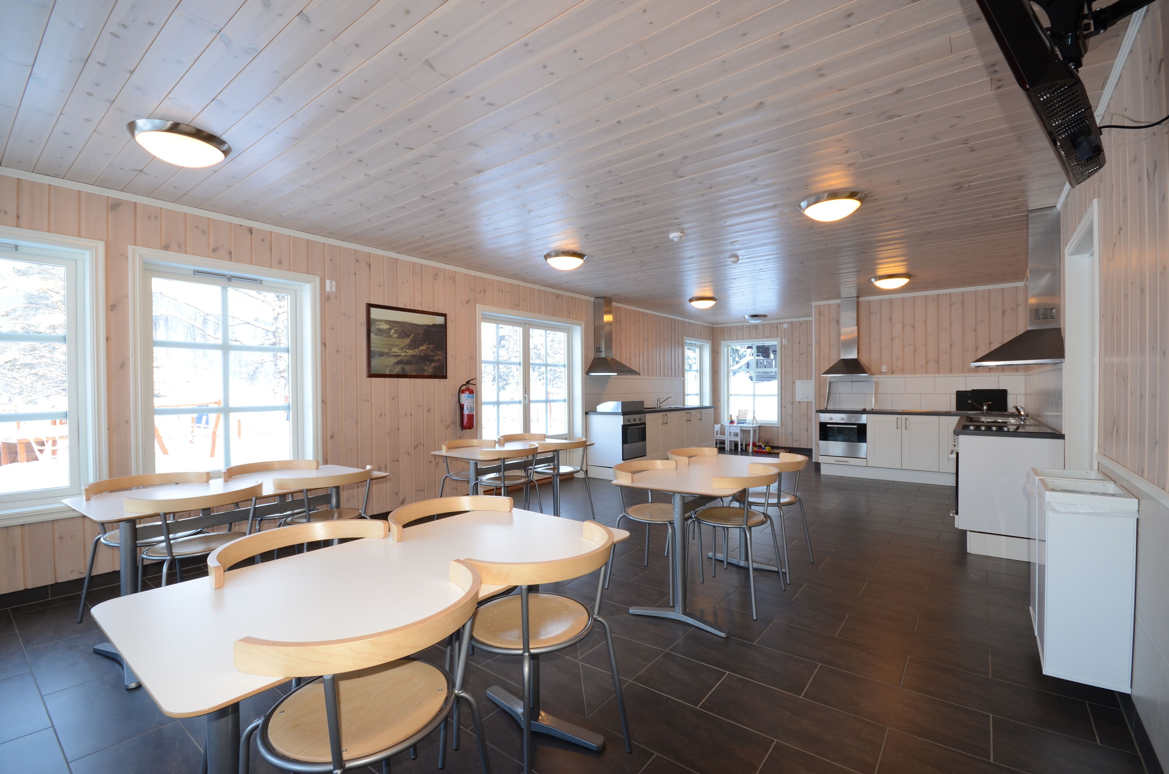 Uma ampla área de refeições com várias mesas e cadeiras, cozinha equipada com eletrodomésticos modernos e grandes janelas que proporcionam iluminação natural.