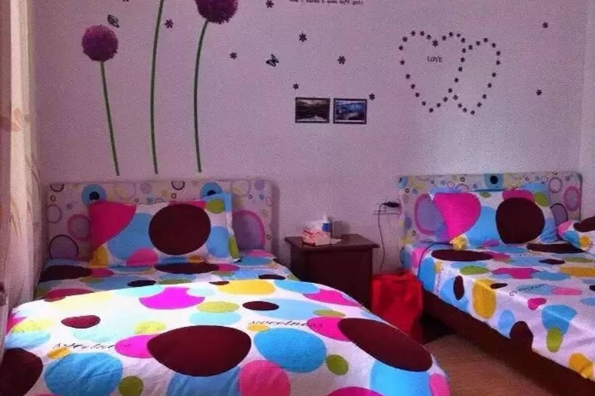 Quarto com duas camas, cada uma com roupa de cama colorida com bolinhas. Decorações nas paredes incluem flores e um coração com a palavra 'LOVE'.