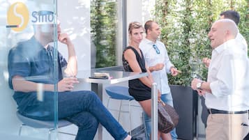 Um grupo de pessoas sentadas e em pé em área externa, separados por uma divisória de vidro, algumas com bebidas na mão.