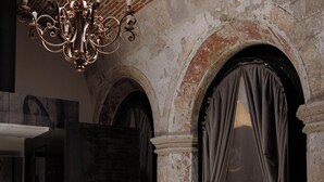 Quarto de hotel com uma cama grande, um candelabro e janelas em arco com cortinas.