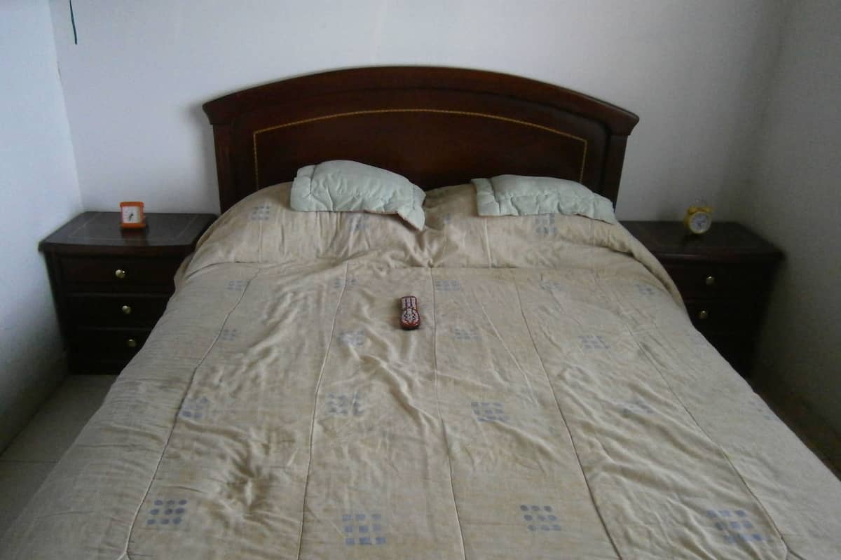 Uma cama com colcha bege e duas almofadas, ladeada por duas mesas de cabeceira de madeira com relógios.