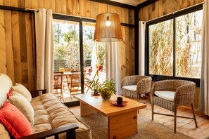 Quarto aconchegante com sofá, duas cadeiras de vime, mesa de madeira com uma planta e janelas amplas com vista para árvores.