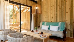 Um quarto aconchegante com piso de madeira, um sofá com manta verde, cadeiras de vime, uma mesa de madeira com uma planta e duas xícaras, e uma janela grande com vista para o exterior.