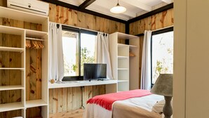 Um quarto com piso de madeira, uma cama com cobertor vermelho, uma escrivaninha com televisão e uma janela com cortinas.