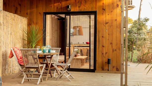 Um deck de madeira com mesa e cadeiras, um lustre pendurado e uma porta de vidro deslizante que dá para um espaço interno.