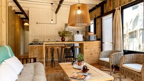 Uma sala de estar aconchegante, com móveis de madeira, um sofá, uma mesa de centro com revistas, cadeiras de vime e uma área de cozinha com banquetas e um lustre.