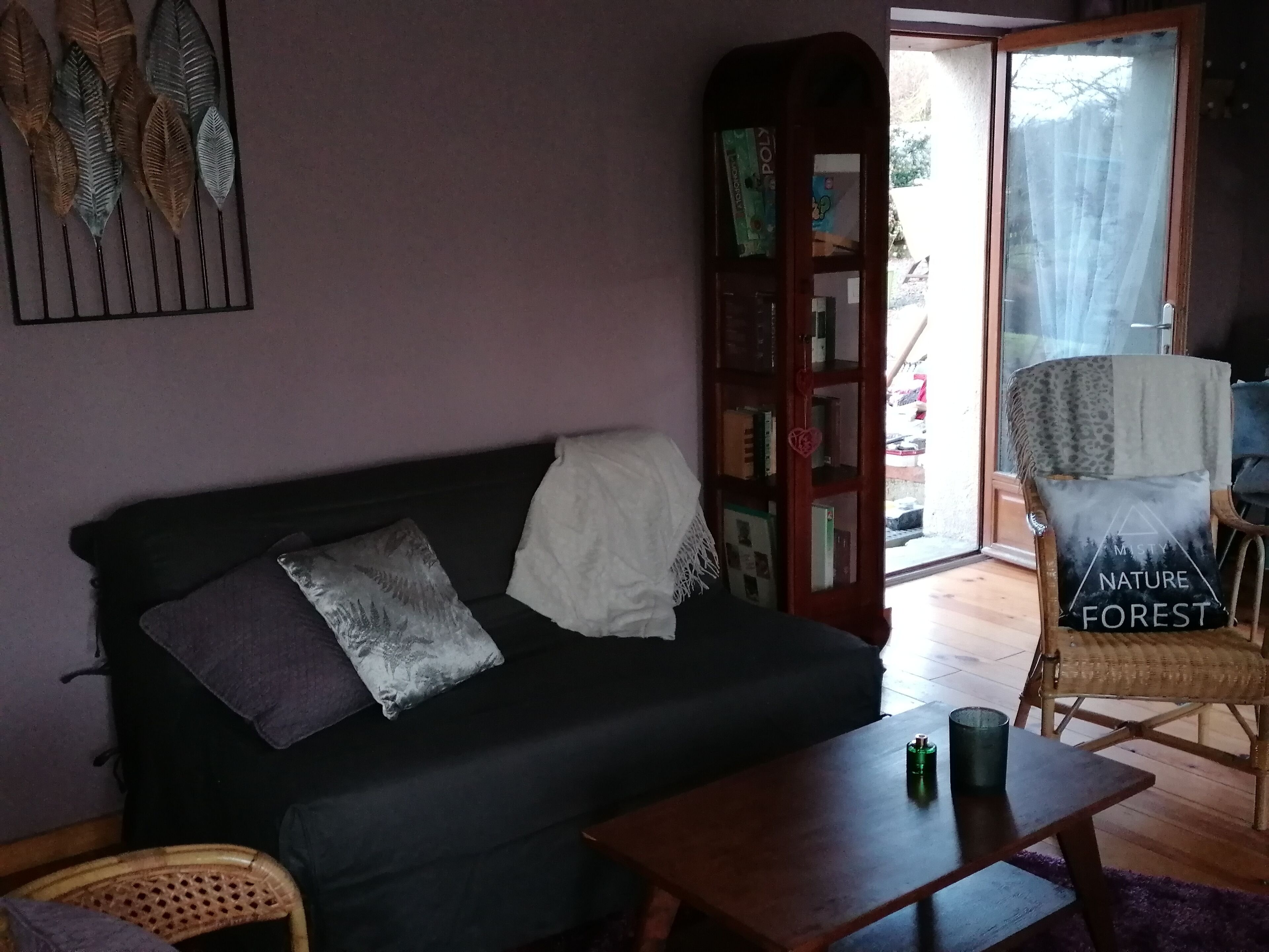 Uma sala de estar com um sofá escuro, uma mesa de centro de madeira, uma cadeira de vime e uma estante com livros e objetos decorativos.