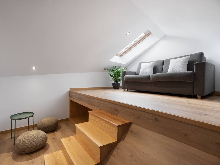 Quarto de loft moderno com teto inclinado, escada de madeira embutida, sofá cinza e uma pequena mesa verde.