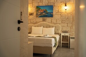 Quarto de hotel com parede de pedra, cama com roupa de cama branca, pintura de uma paisagem costeira e mesa de cabeceira com abajur.