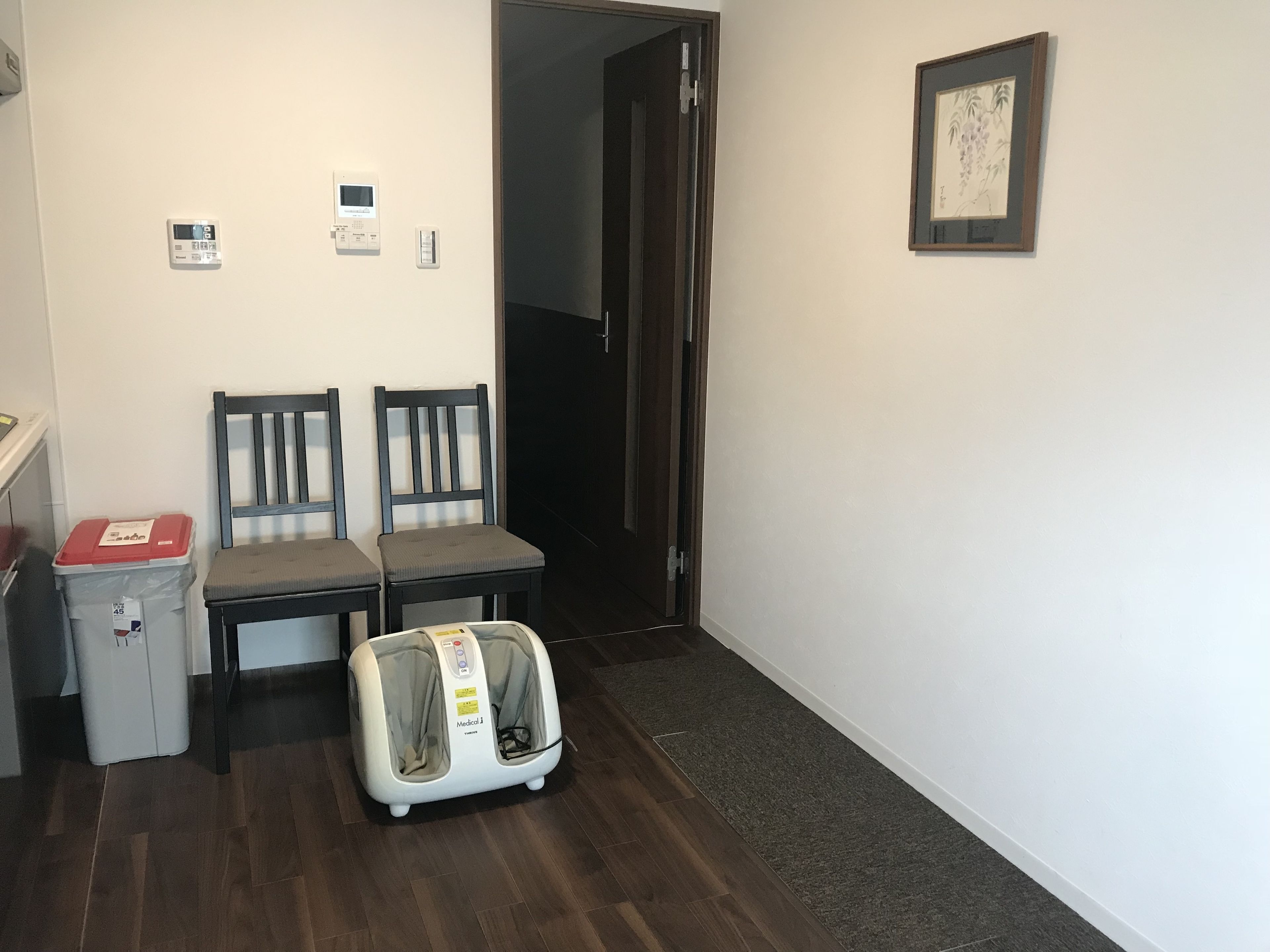 Um corredor com duas cadeiras de madeira, uma mala branca, um cesto de lixo e uma porta.