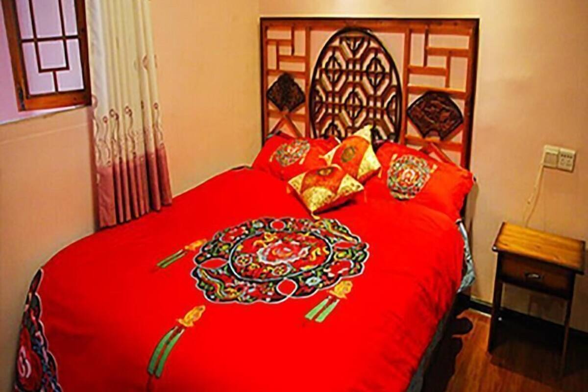 Cama com colcha vermelha e almofadas decorativas, cabeceira de madeira, mesa de cabeceira e janela com cortinas.