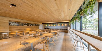Um café moderno com mesas e cadeiras de madeira, área de cozinha e janelas amplas com plantas.