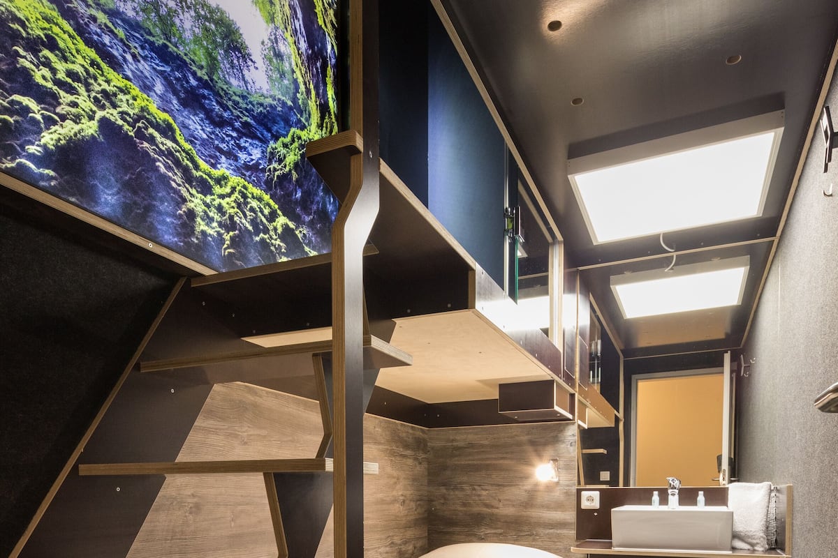 Quarto de hotel compacto com beliches, uma kitchenette pequena e uma janela grande que traz luz natural.