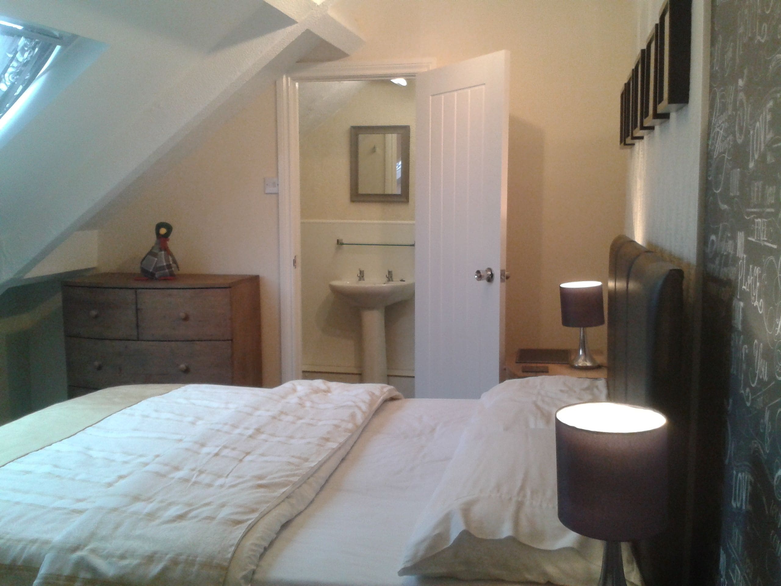 Quarto com cama, cômoda, abajur e banheiro visível através de uma porta aberta.