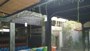 Uma área externa coberta com piso de azulejos, um banco com almofadas, plantas pendentes e um vaso com planta.