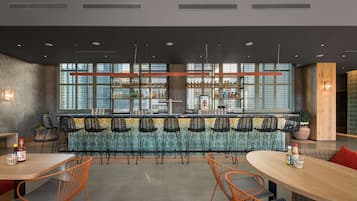 Uma área de bar moderna com um balcão extenso, diversas opções de assento e vista para a cidade através de grandes janelas.
