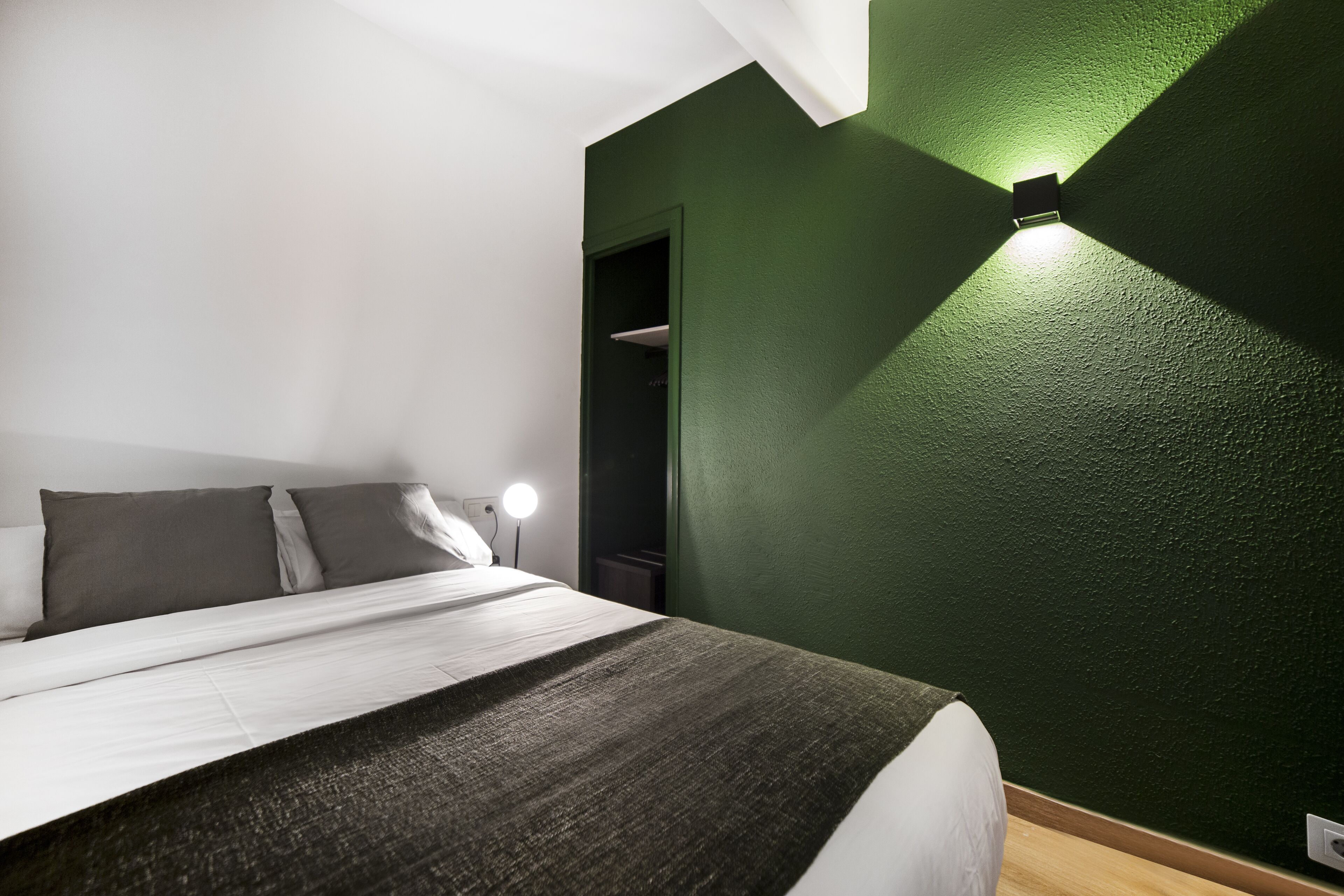 Quarto de hotel moderno com cama bem arrumada, parede de destaque verde e pequena mesa de cabeceira com abajur.