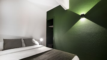 Quarto de hotel moderno com cama bem arrumada, parede de destaque verde e pequena mesa de cabeceira com abajur.