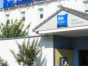 Um edifício com a sinalização 'ibis budget hotels', um pequeno caixa eletrônico e uma árvore na frente.