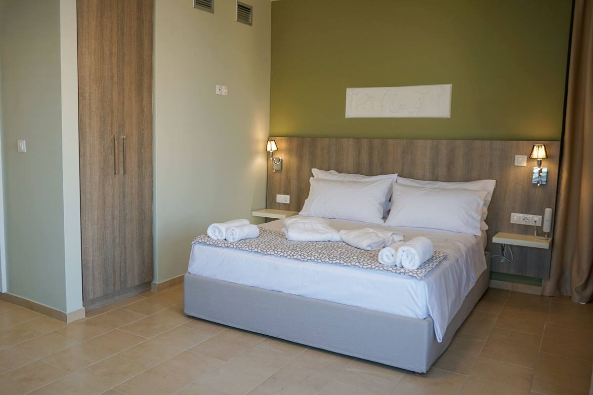 Quarto de hotel com cama, duas mesas de cabeceira, um criado-mudo, luminária na parede e porta-toalhas.
