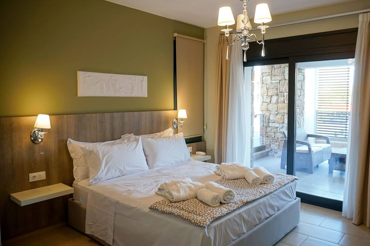 Quarto de hotel com uma cama grande, duas luminárias de cabeceira, uma mesa de cabeceira, uma obra de arte na parede e uma porta de correr de vidro que dá para uma área externa com mobiliário.