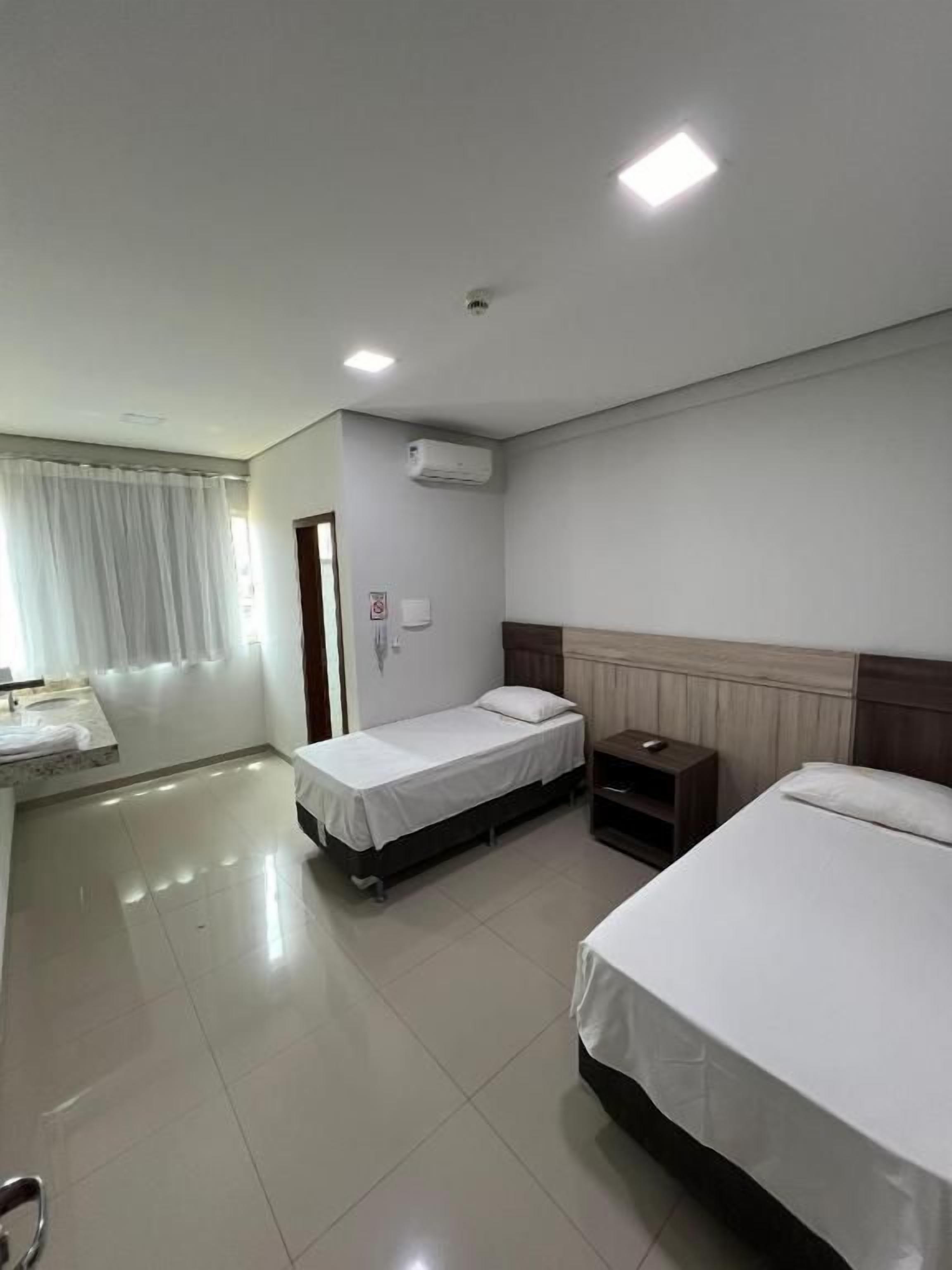 Quarto de hotel com duas camas, uma pequena mesa de cabeceira, um criado-mudo, uma janela com cortinas, ar condicionado fixado na parede e piso em azulejos.