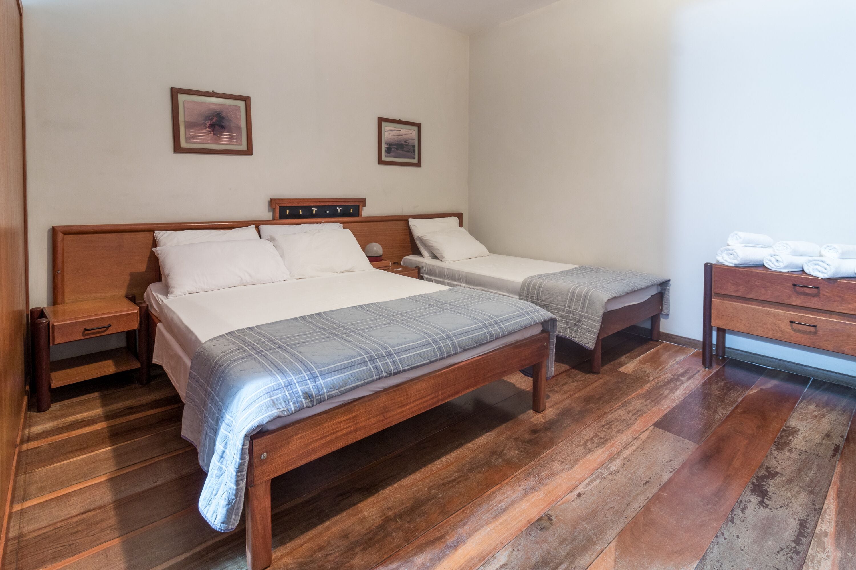 Quarto com duas camas, piso de madeira, mesa de cabeceira, cômoda e duas imagens emolduradas na parede.