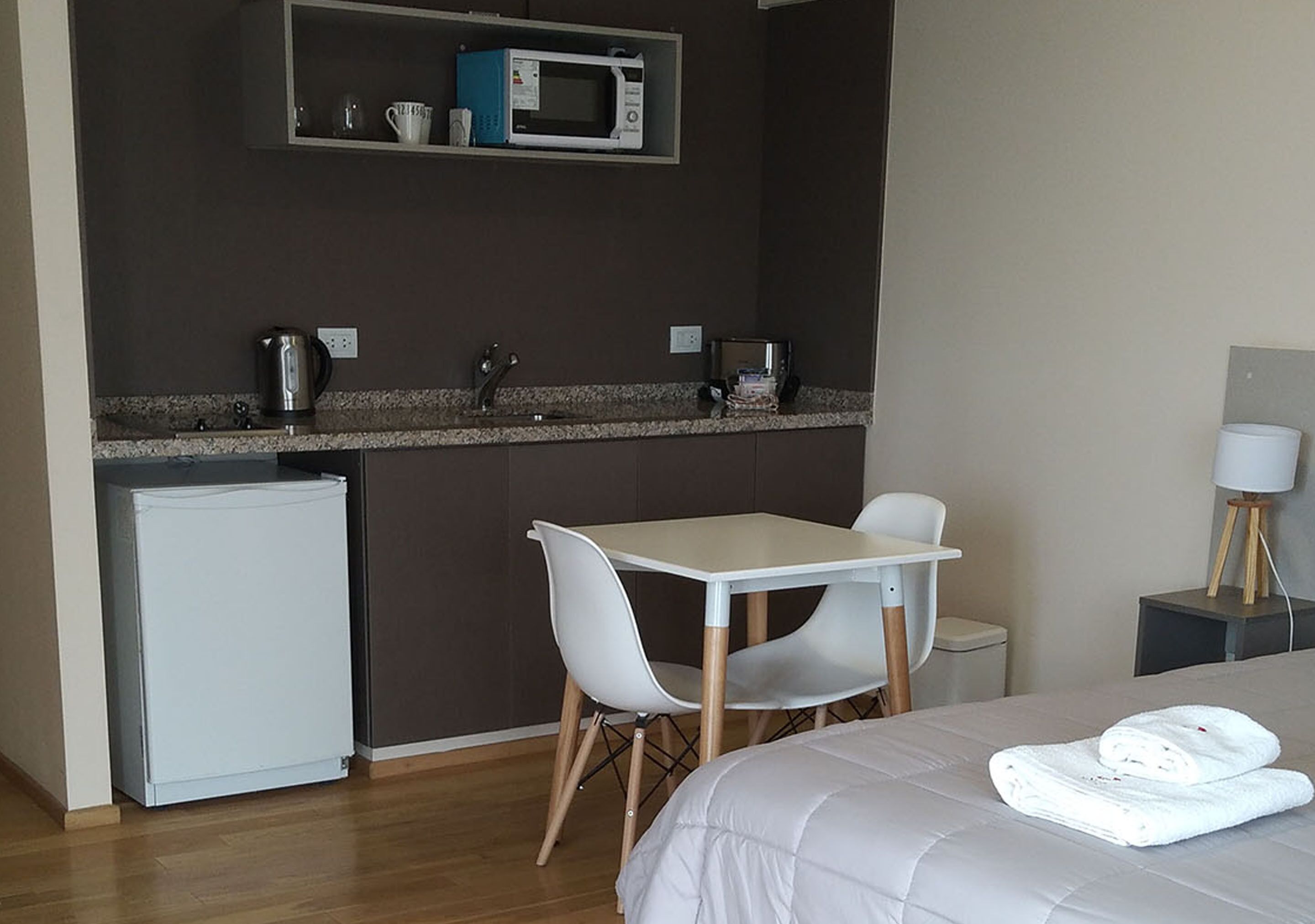 Quarto de hotel compacto com cozinha compacta, mini-geladeira, micro-ondas, cafeteira, mesa para dois, cama com edredom branco e luminária de cabeceira.