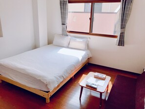 Quarto individual com uma cama de madeira, roupa de cama branca, uma mesinha com um controle remoto e uma janela com cortinas.