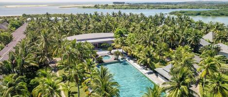 Um resort com piscina central rodeada por palmeiras, um edifício grande e vista para o mar.