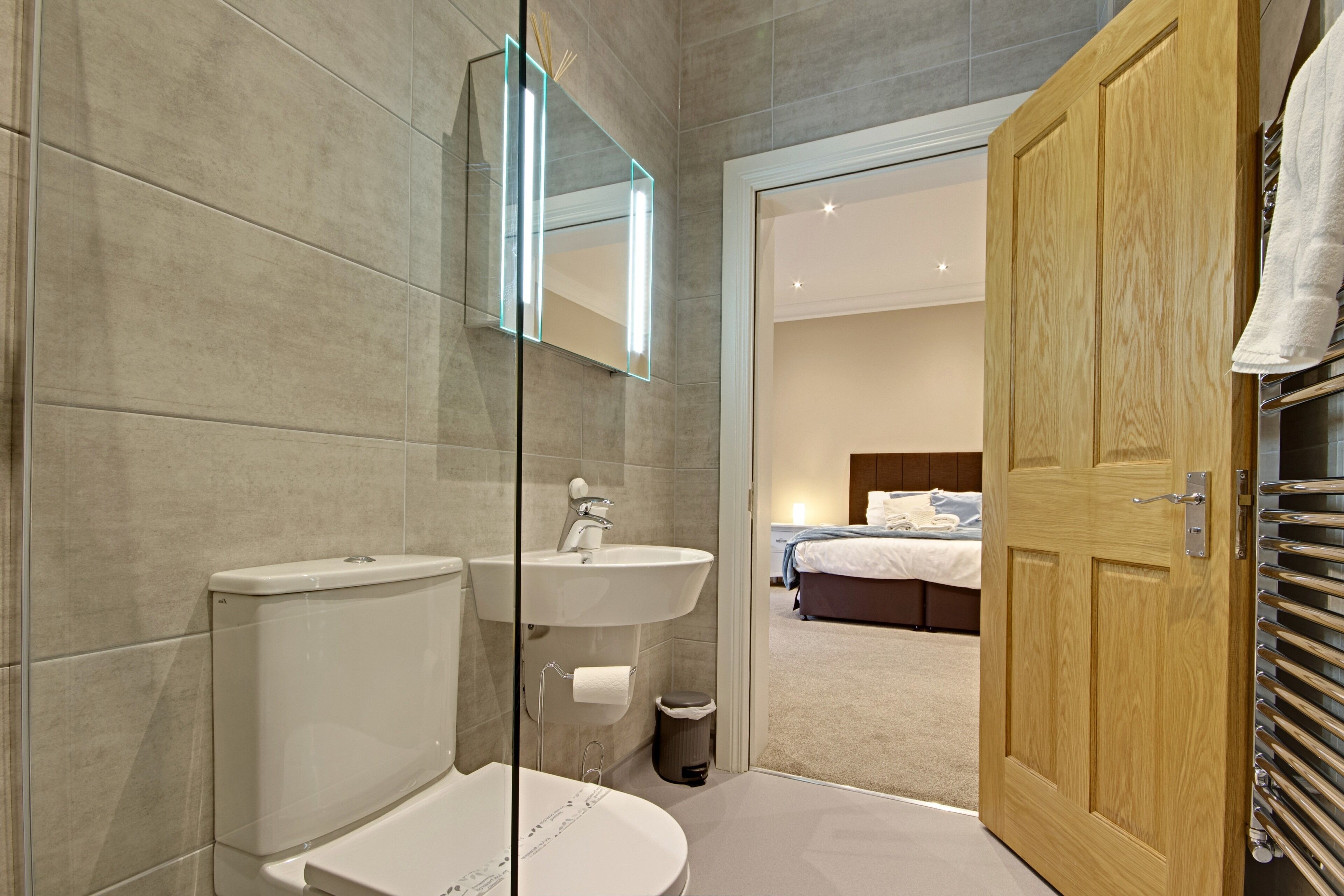 Uma casa de banho moderna com sanita, lavatório e espelho. Uma porta de madeira dá acesso a um quarto com cama e mesa de cabeceira.