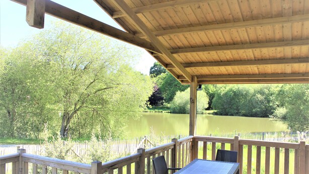 Um deck de madeira com mesa e cadeiras oferece vista para um lago, rodeado por árvores e um cercado de madeira.