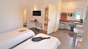 Quarto de hotel compacto com duas camas, um pequeno espaço de cozinha, uma televisão e um banheiro.