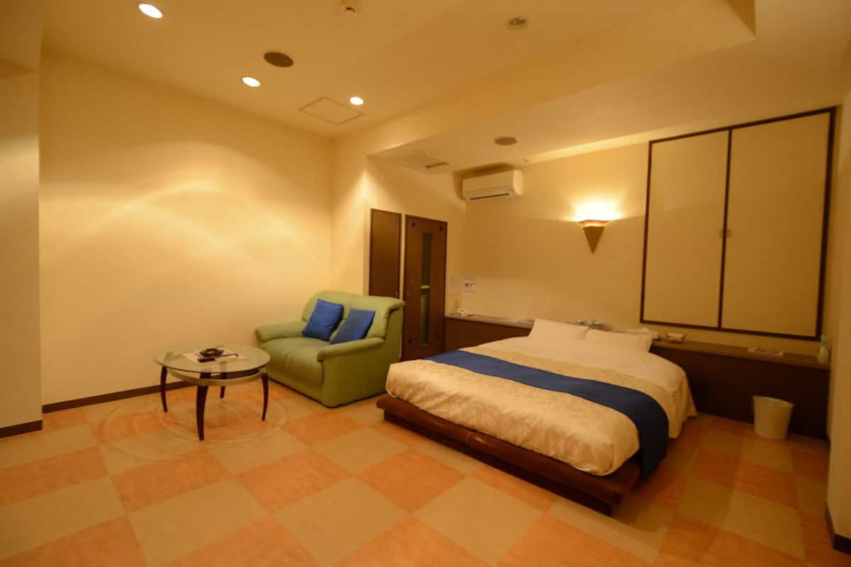 Quarto de hotel com cama, poltrona verde, mesa pequena e espelho grande.