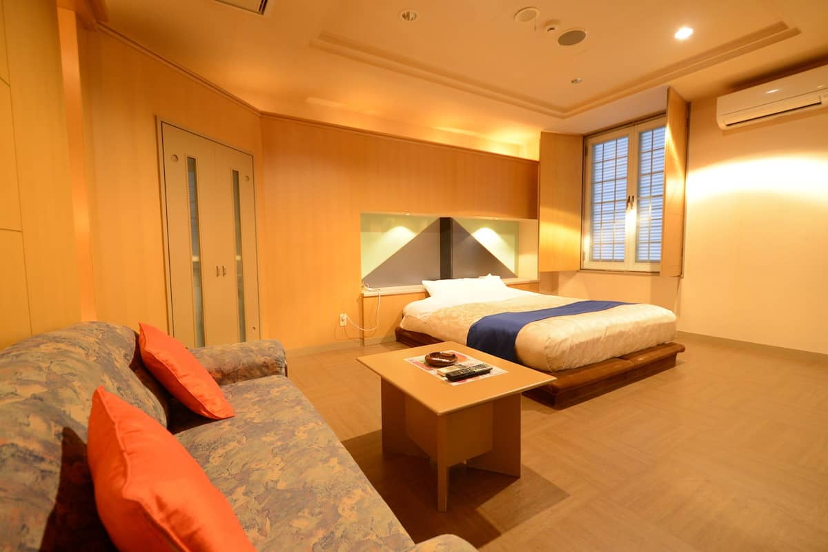 Quarto de hotel com cama, sofá, mesa pequena e janela com persianas.