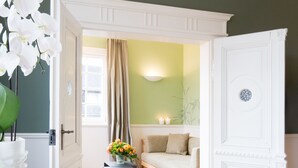 Um quarto com piso de madeira em padrão herringbone, porta branca, sofá bege e uma pequena mesa com um vaso de flores.
